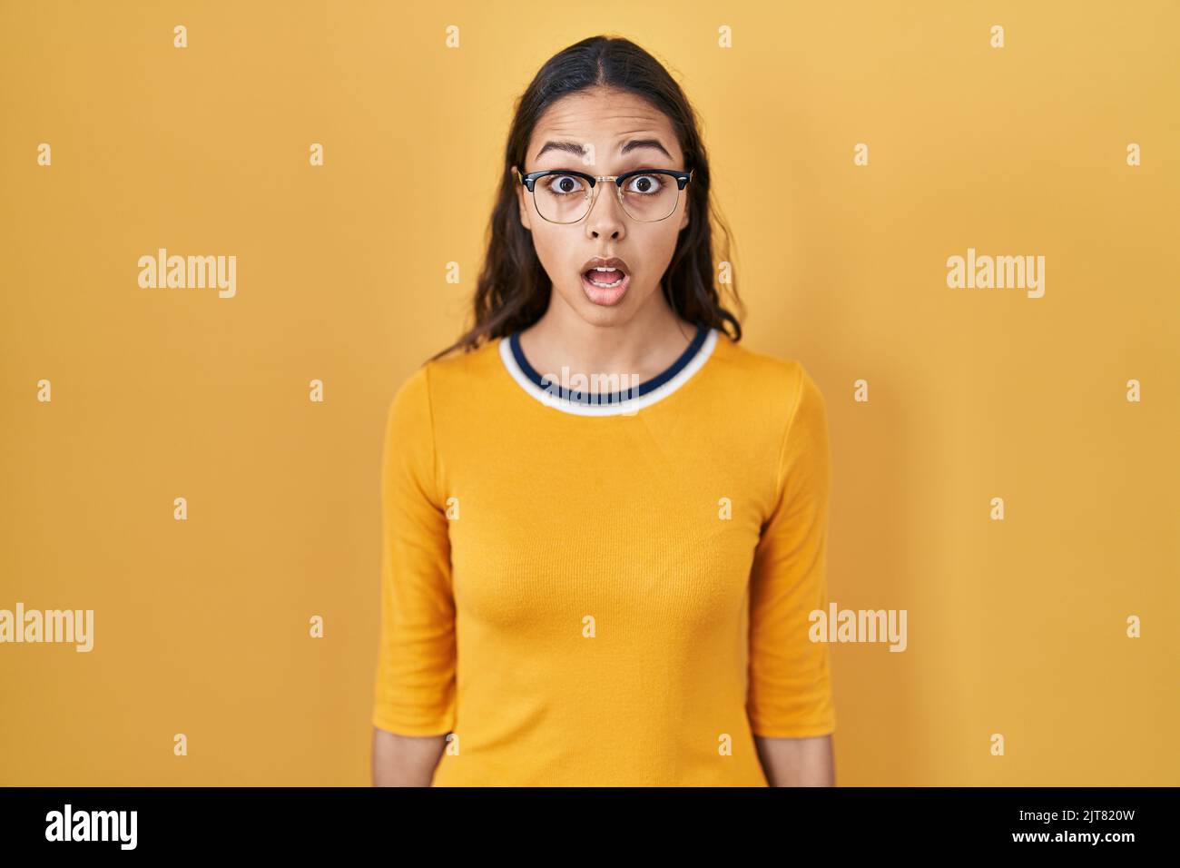 Young brazilian woman wearing glasses over yellow background scared and ...