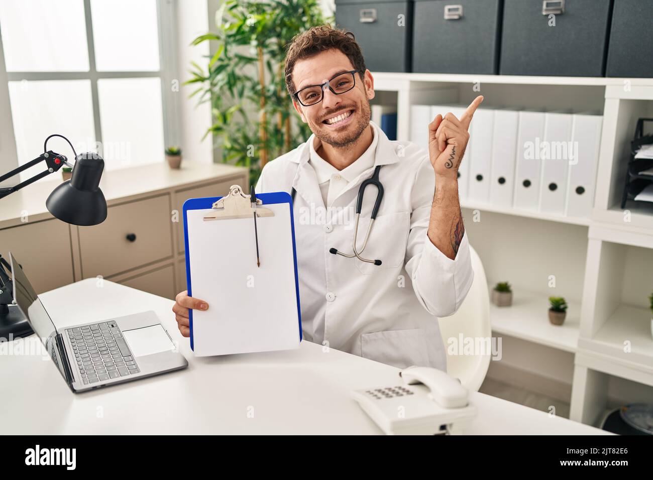 Young hispanic man wearing doctor stethoscope holding clipboard smiling ...