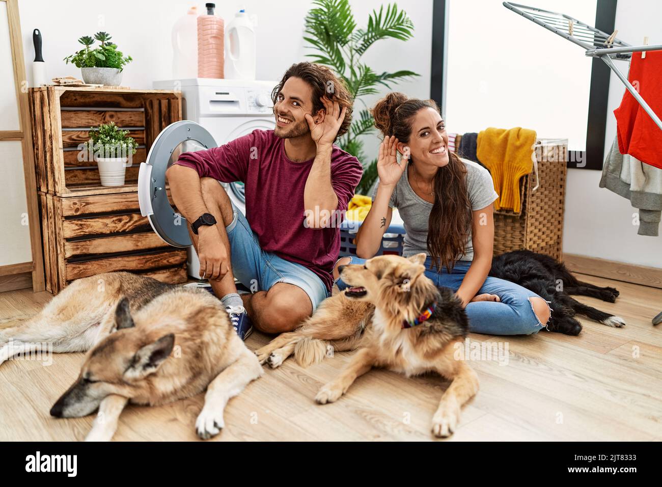 Young hispanic couple doing laundry with dogs smiling with hand over ...