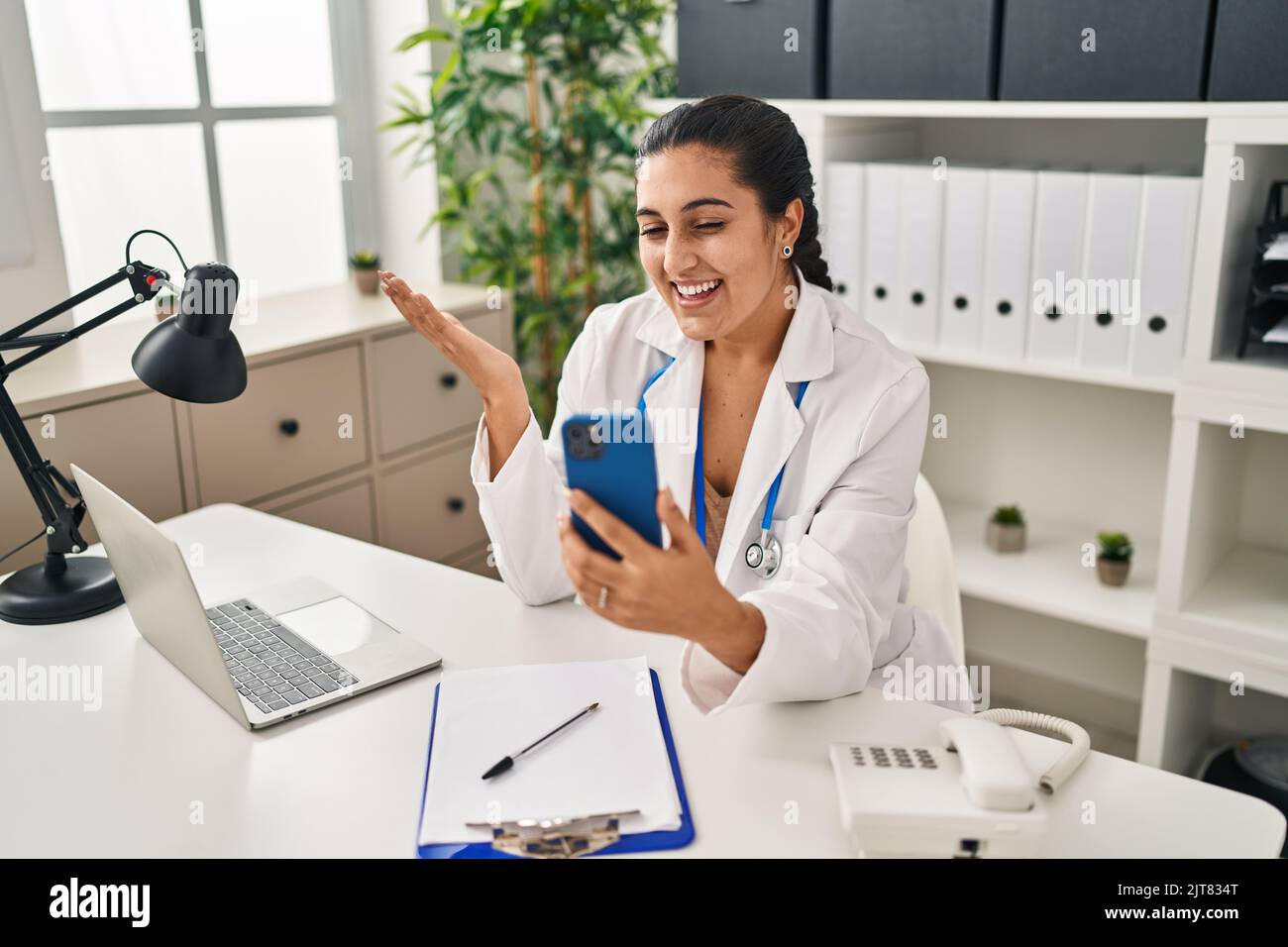Young doctor hispanic woman doing video call celebrating achievement with happy smile and winner ...