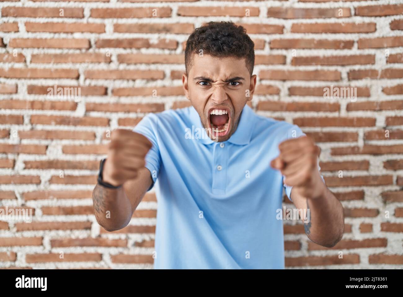 Brazilian young man standing over brick wall angry and mad raising ...