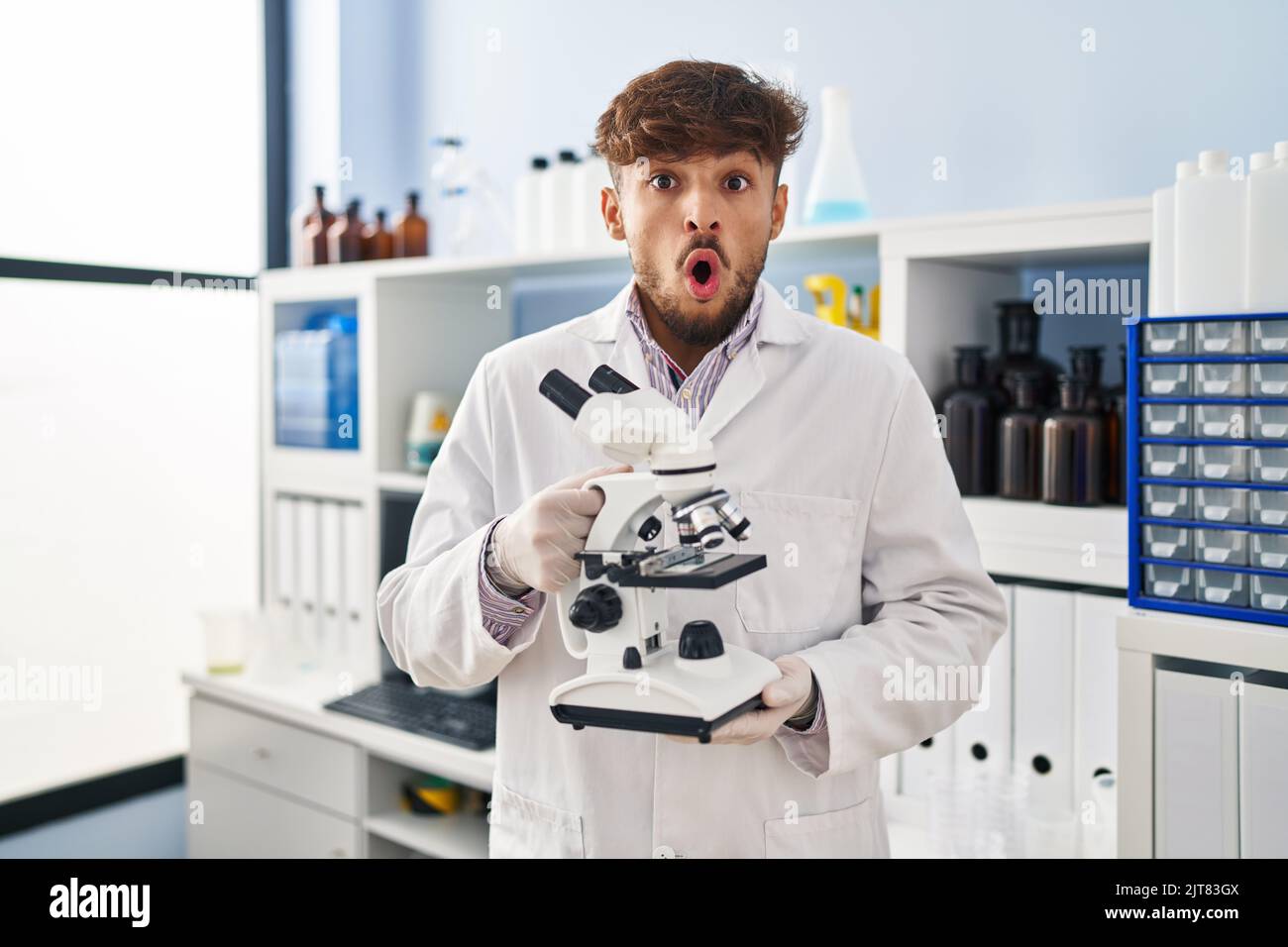 Arab man with beard working at scientist laboratory holding microscope in shock face, looking ...