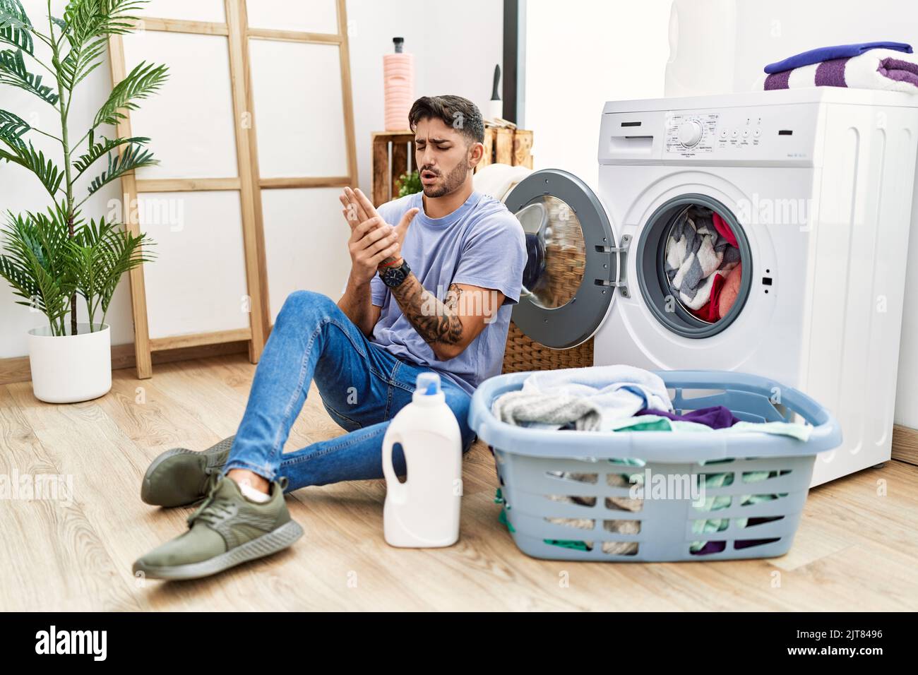 Young hispanic man putting dirty laundry into washing machine suffering ...