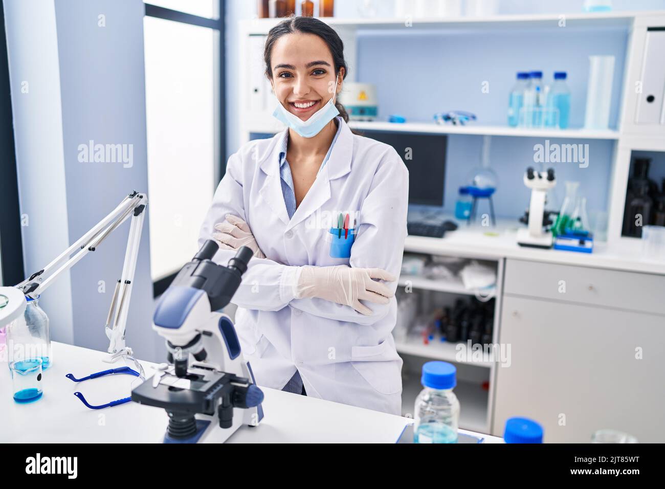 Young hispanic woman wearing scientist uniform standing with arms crossed gesture at laboratory ...