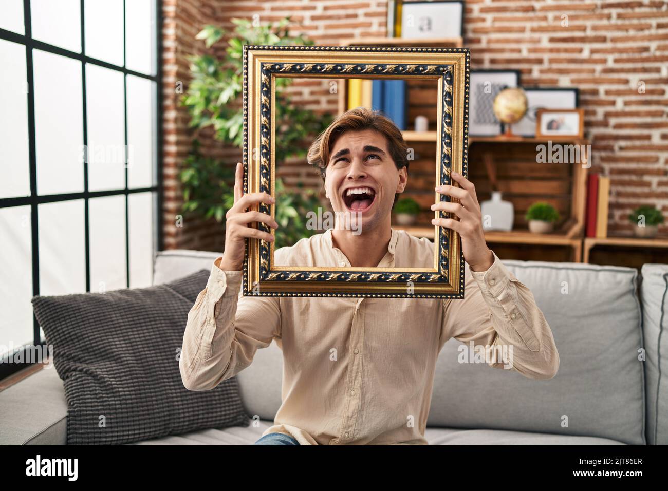 Young man holding empty frame angry and mad screaming frustrated and ...