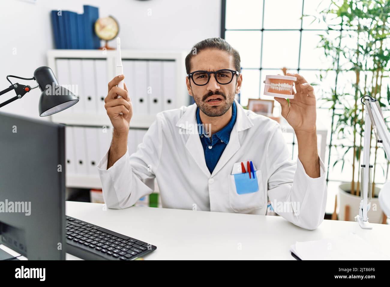 Young hispanic dentist man holding denture and toothbrush clueless and ...
