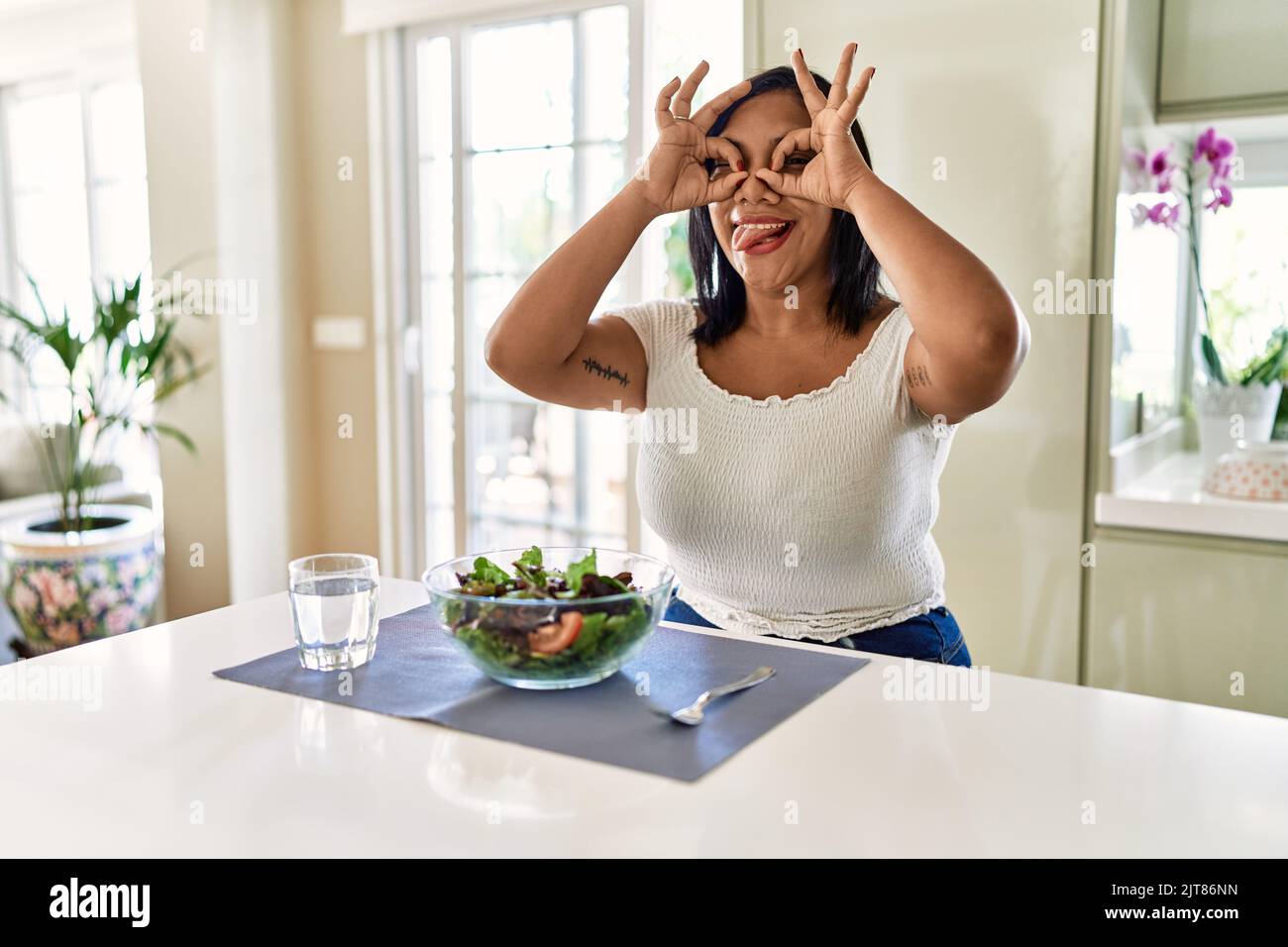 Young hispanic woman eating healthy salad at home doing ok gesture like ...