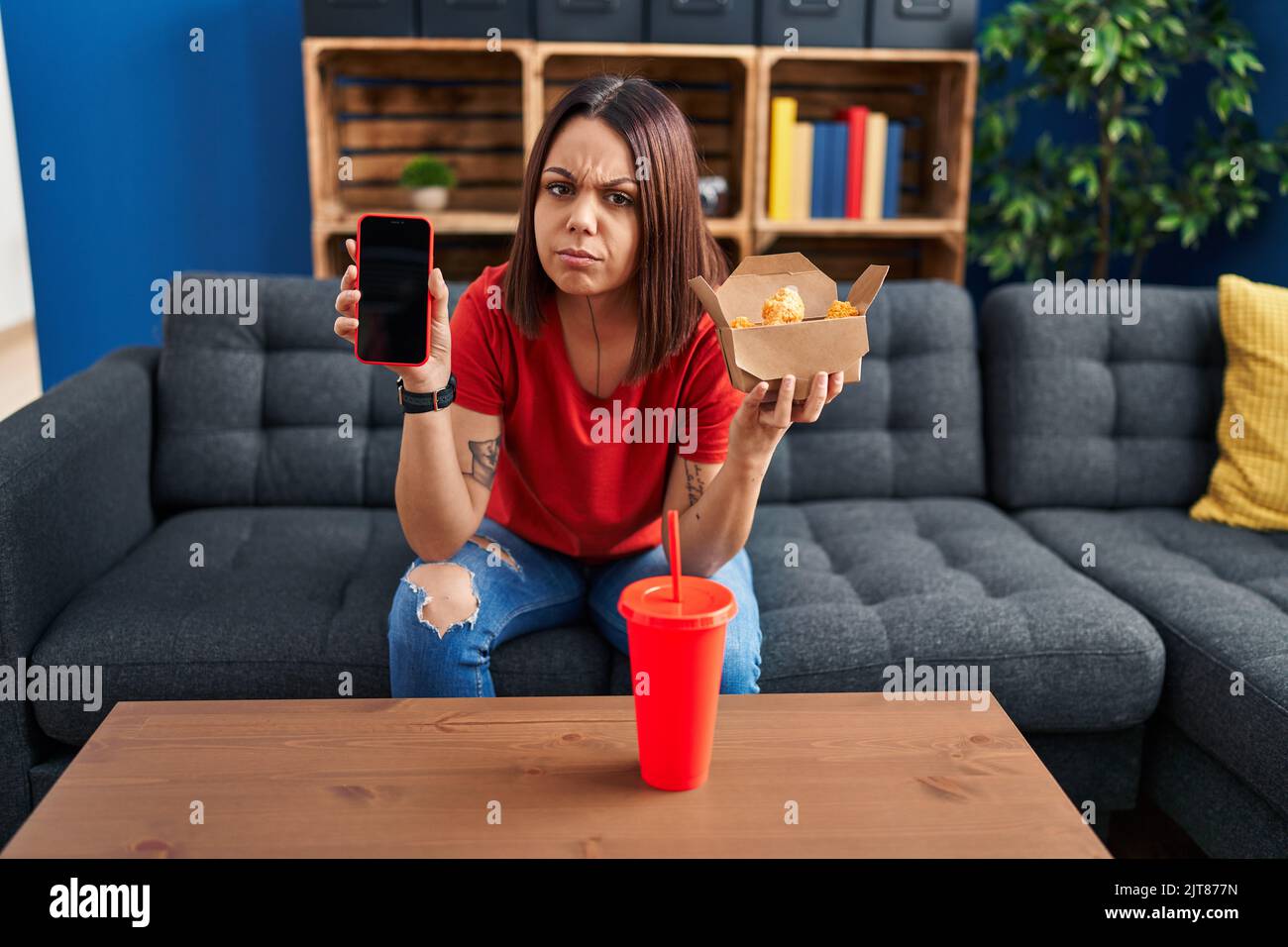 Young hispanic woman eating fast food showing smartphone screen ...