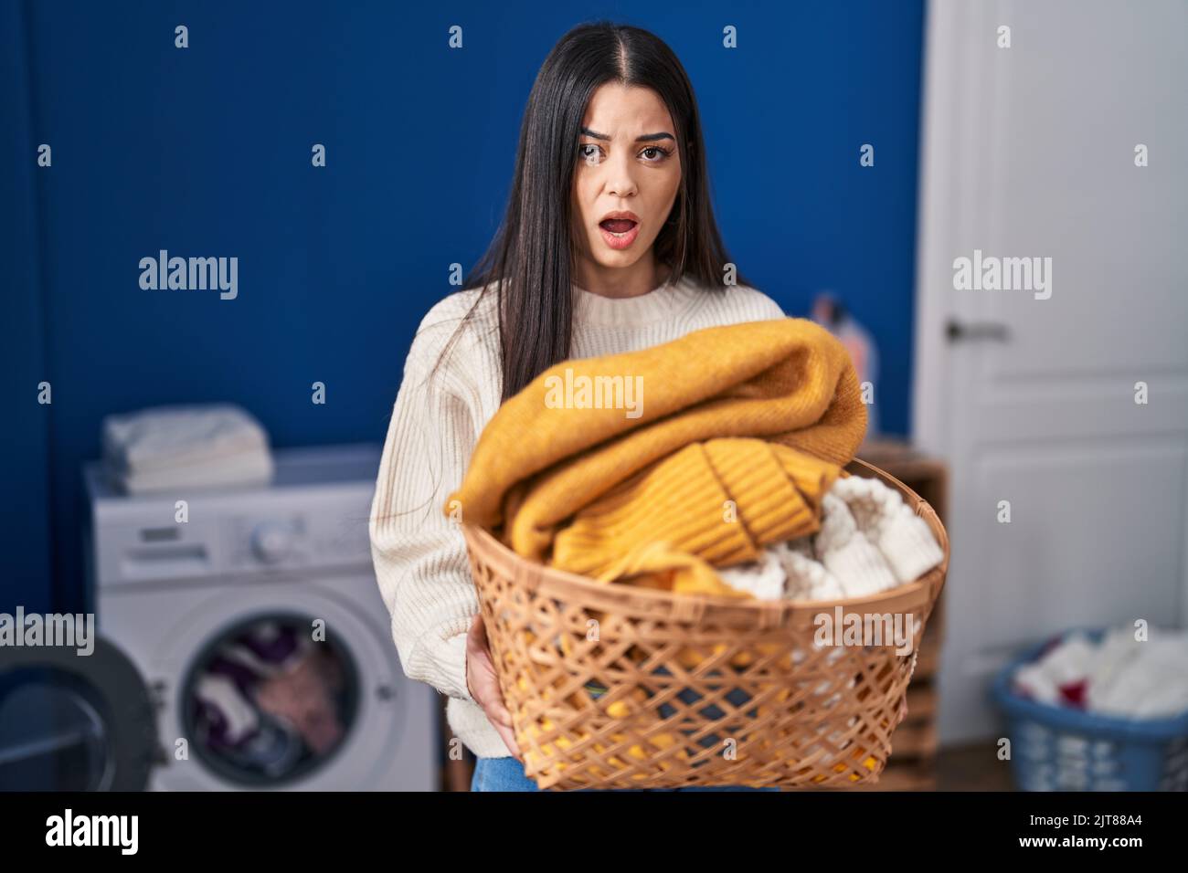 Young woman holding laundry basket in shock face, looking skeptical and ...