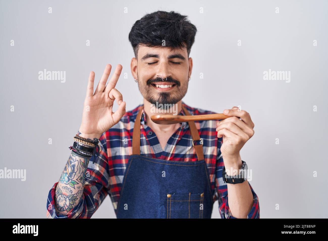 Young hispanic man with beard wearing apron tasting food holding wooden ...