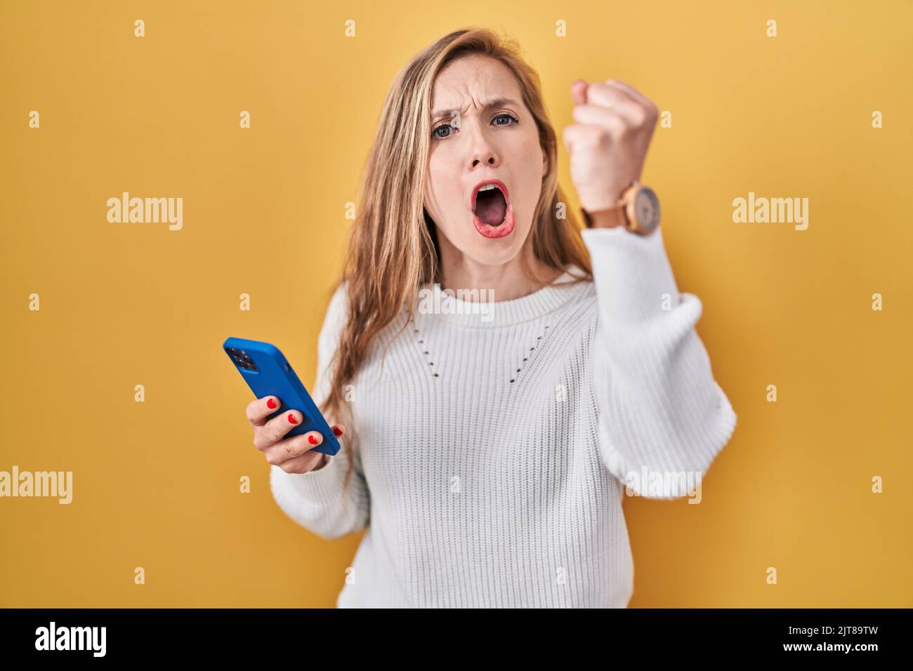 Young blonde woman using smartphone typing message angry and mad ...