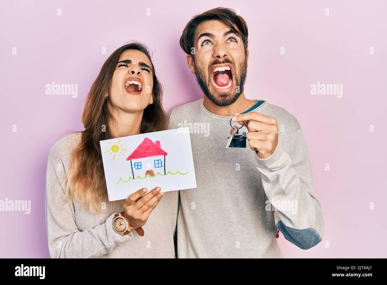Young hispanic couple holding house draw and key of new home angry and ...