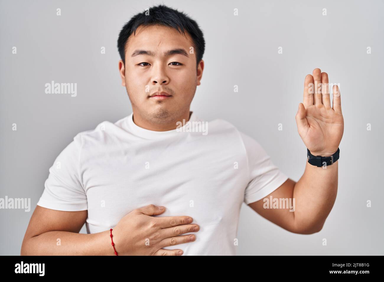 Young chinese man standing over white background swearing with hand on ...