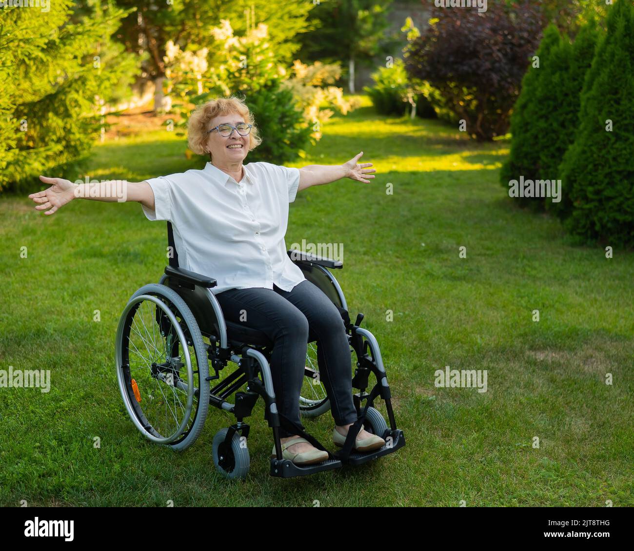 Happy Old Woman Spread Her Arms To The Sides While Sitting In A Wheelchair On A Walk Outdoors