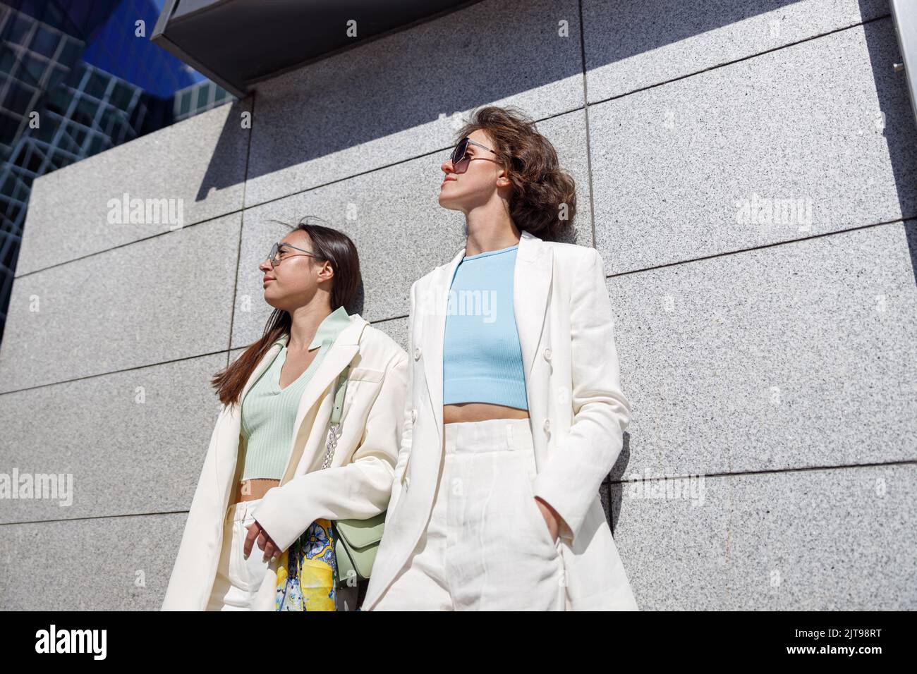 Two woman friends have a rest after walking in city standing near wall ...