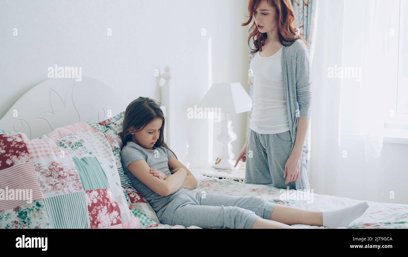 Angry mother argue scolding her upset little daughter lying on bed in cozy bright bedroom at ...