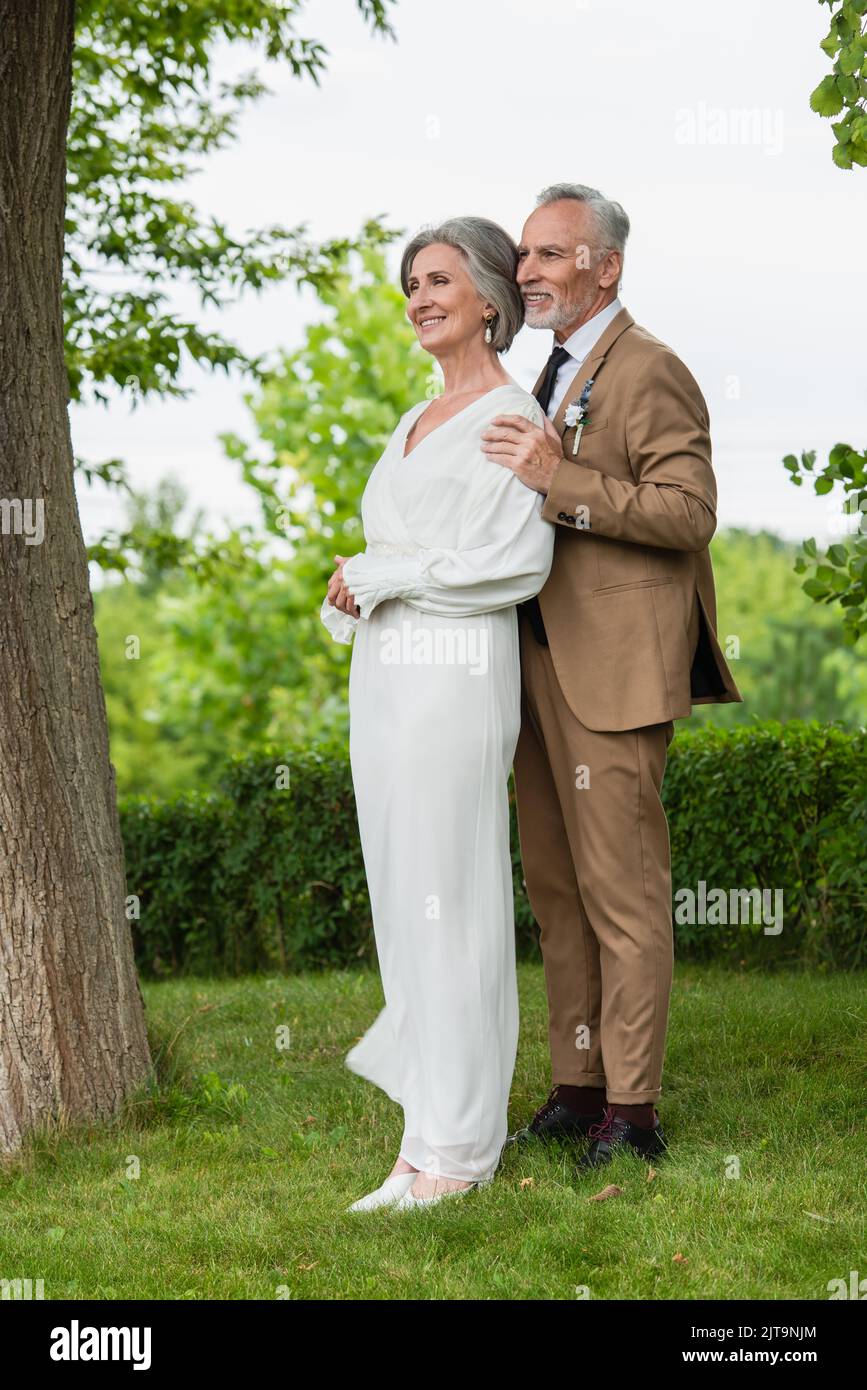 full length of happy middle aged groom in suit hugging mature bride in ...