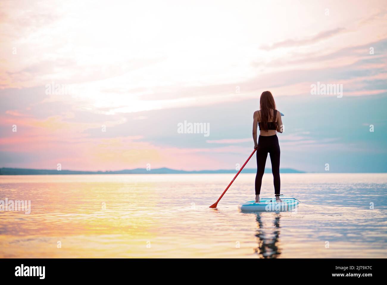 Rear view of girl surfer paddling on surfboard on the lake at sunrise ...