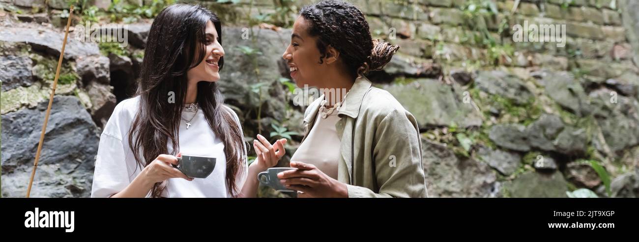 Side view of cheerful multiethnic friends with cups talking in outdoor ...