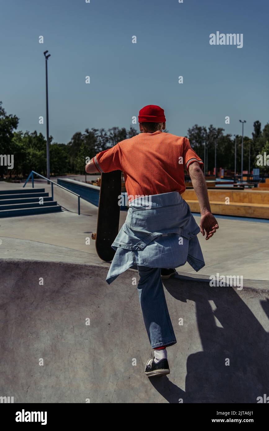 back view of man in orange t-shirt running on ramp with skateboard ...
