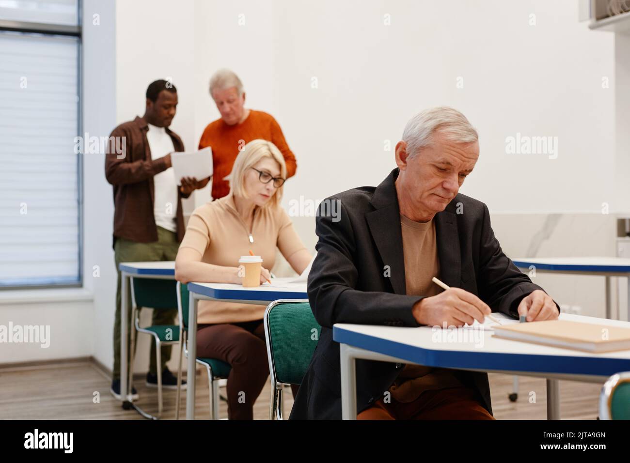 Senior people sitting at desks in a row and writing test at classroom ...