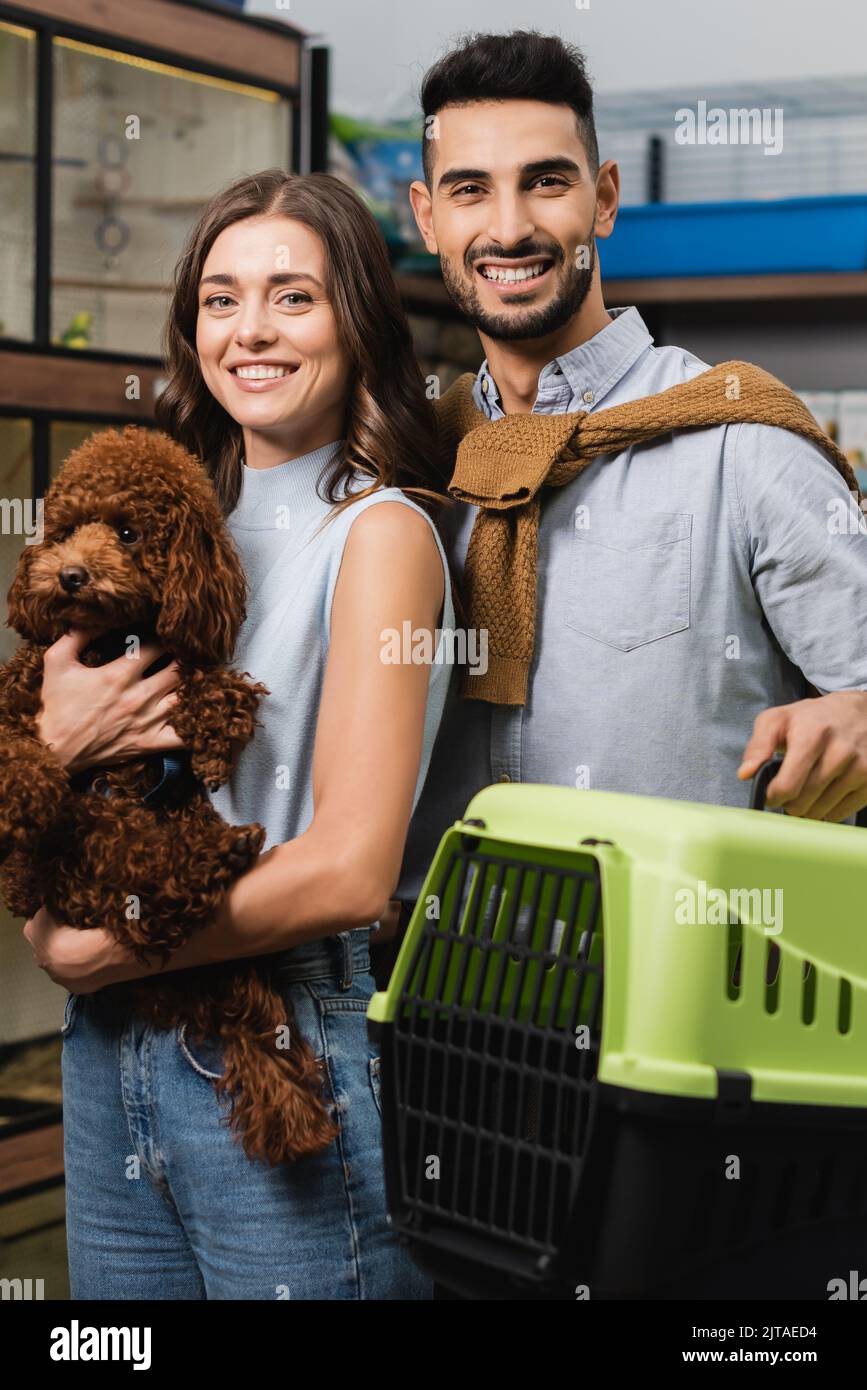 Cheerful multiethnic couple holding poodle and animal cage in pet shop ...