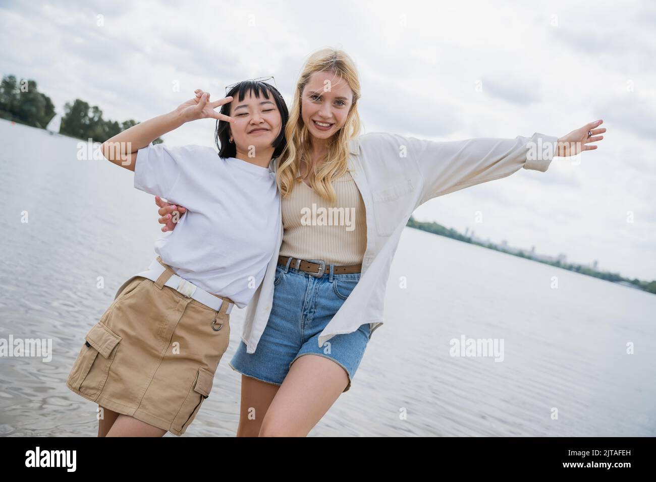 happy blonde woman embracing asian friend showing victory sign while ...