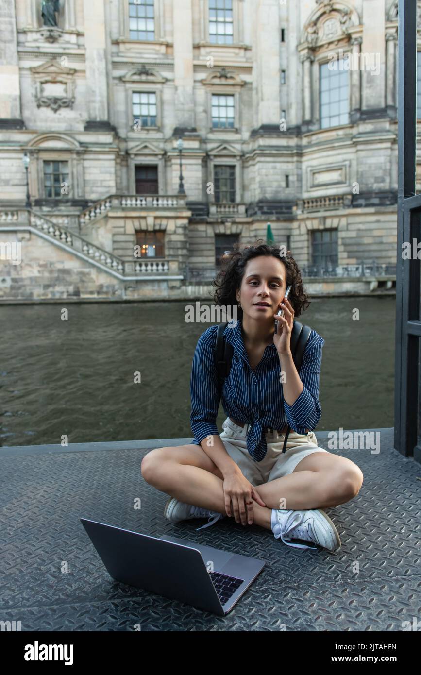 curly woman talking on smartphone while sitting with crossed legs near ...