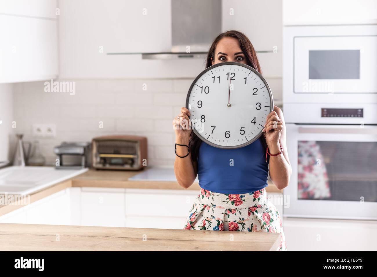 Woman stands in the kitchen holding big clock showing exactly twelve o ...
