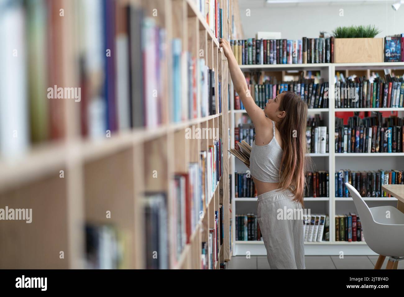 A Caucasian teenage girl chooses a book among the bookshelves in a public library. Background ...