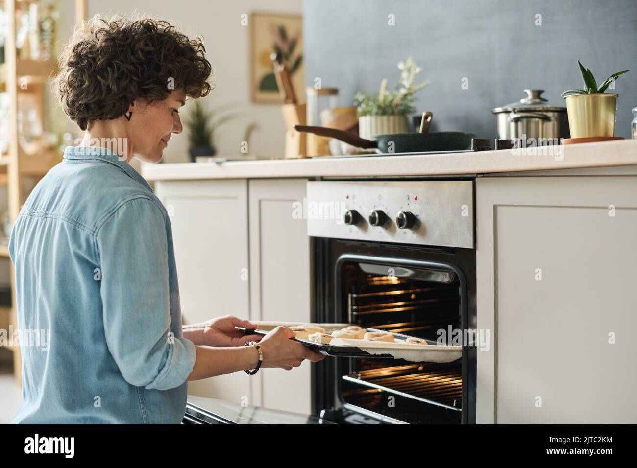 Young woman putting tray with buns in the oven while cooking in ...