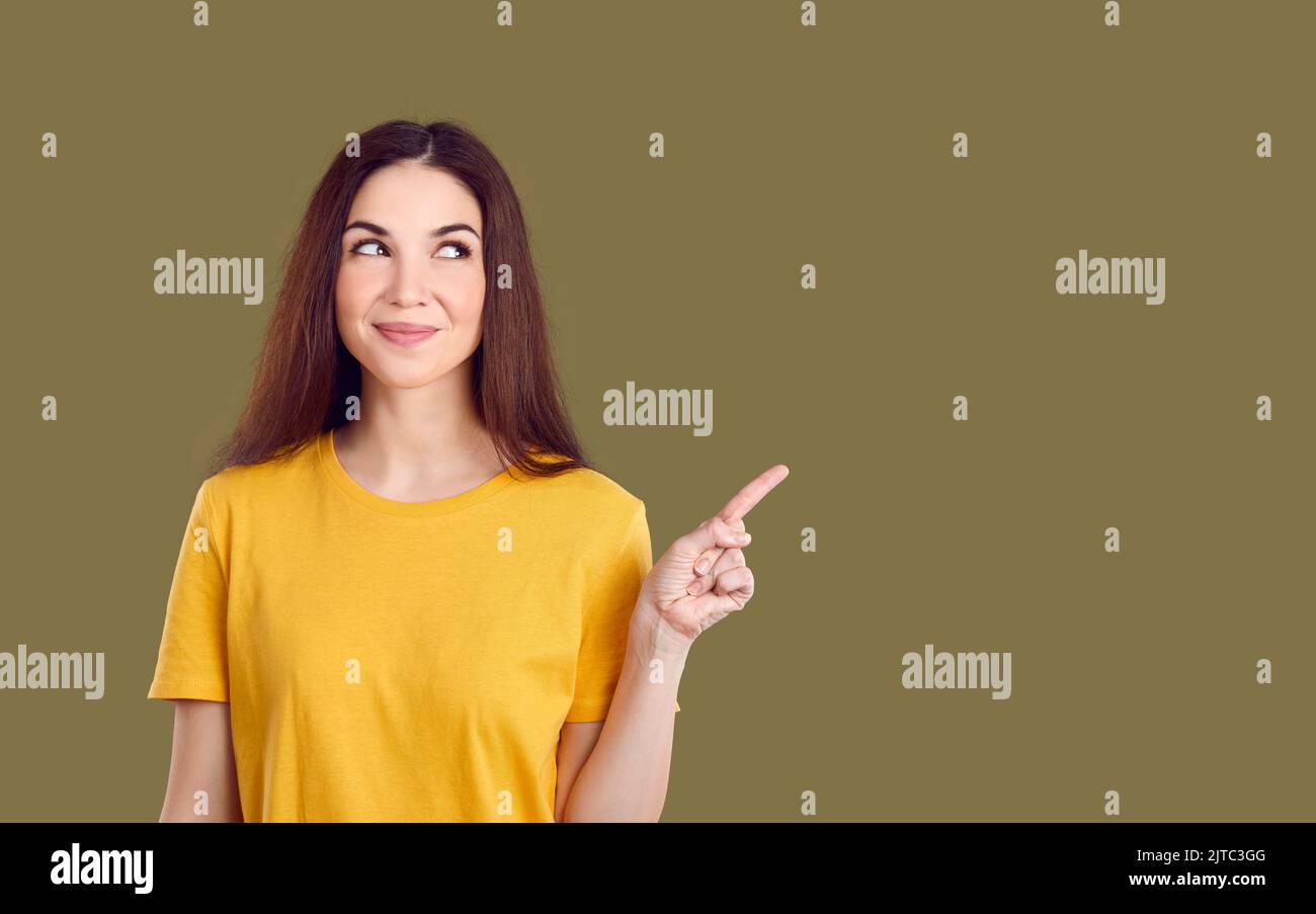 Smiling young woman points finger and flirtatiously looks at copy space ...