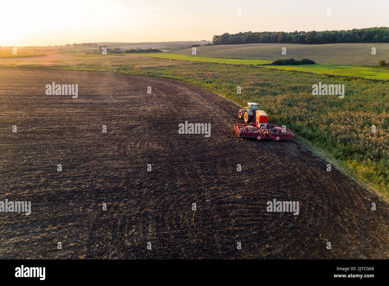 Tractor with sowing machine driving through brown field with greenery ...