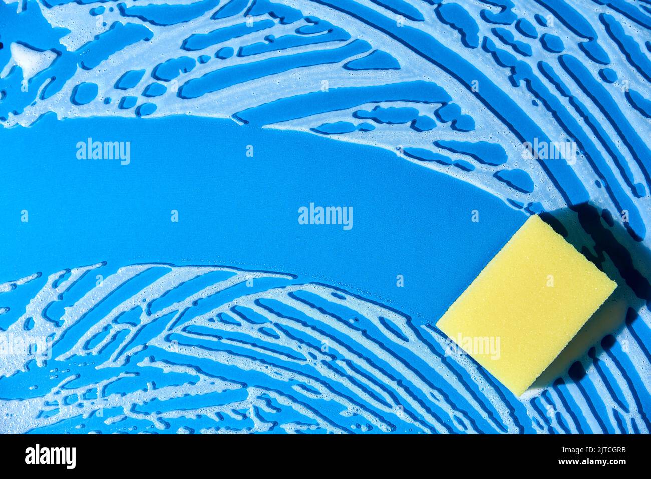Yellow dishwashing sponge on a blue glass surface covered with soap ...