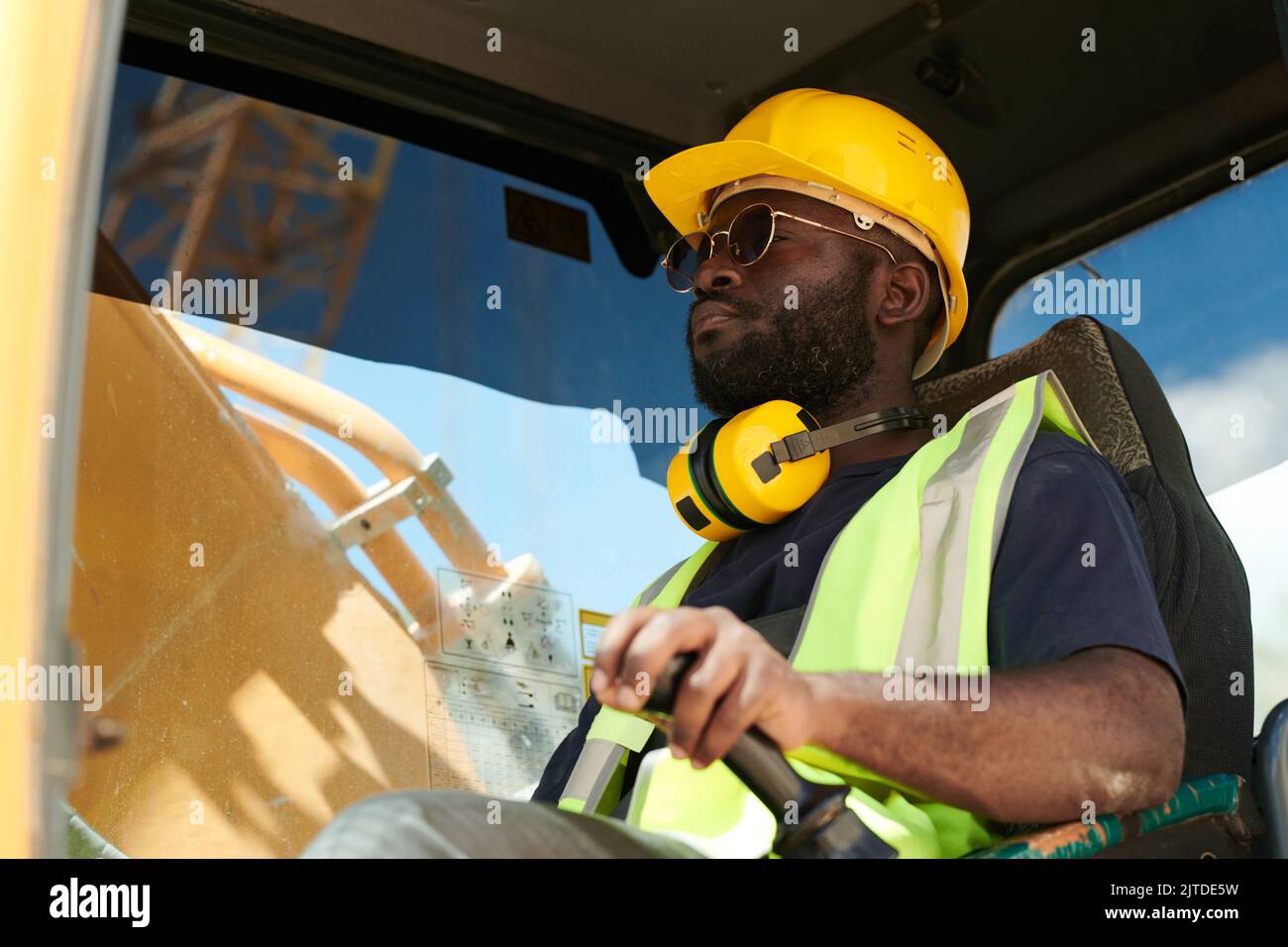 Young black male driver of construction machine in safety helmet and ...