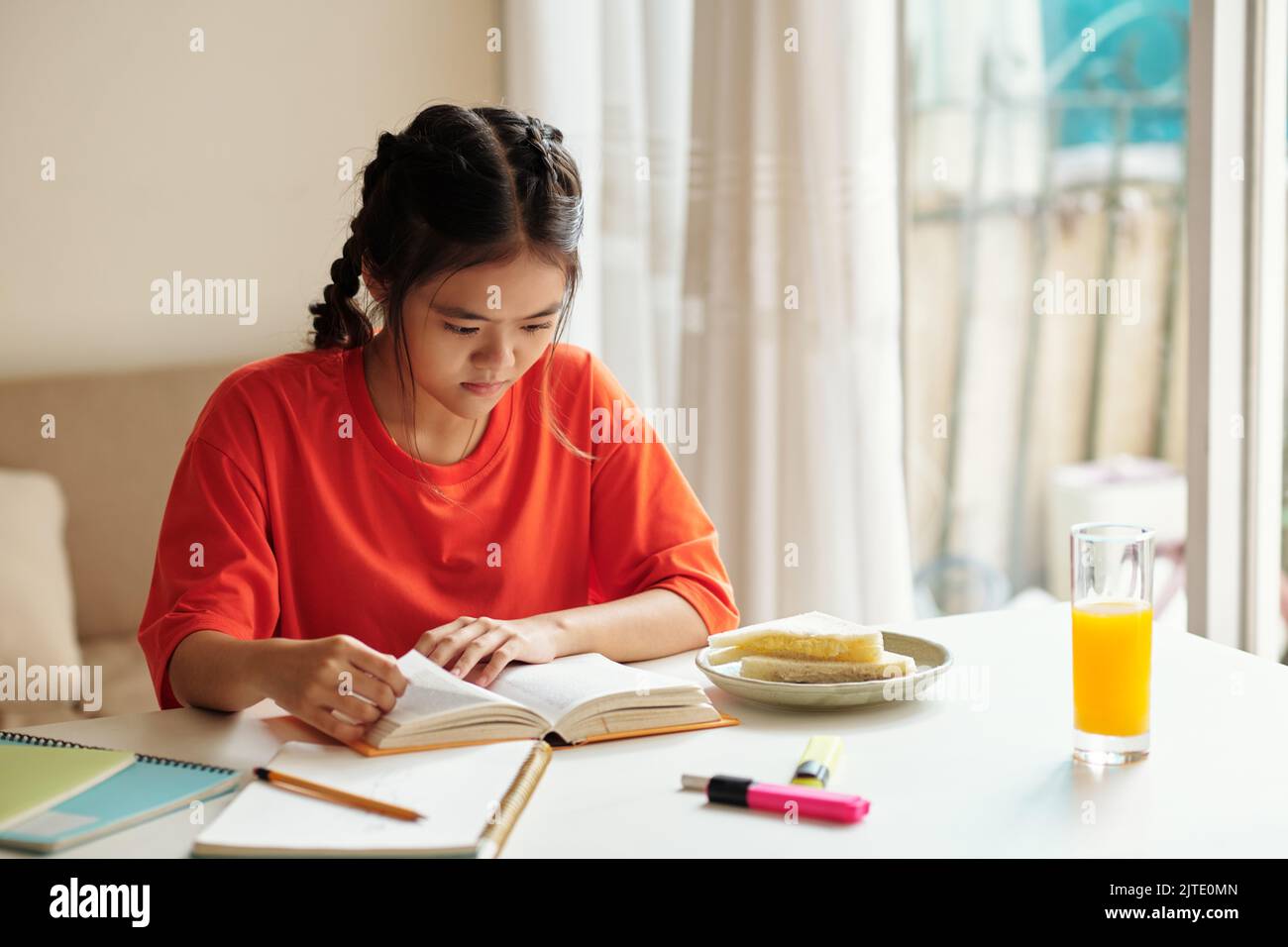 Confused schoolgirl having sandwich and glass of juice when reading ...