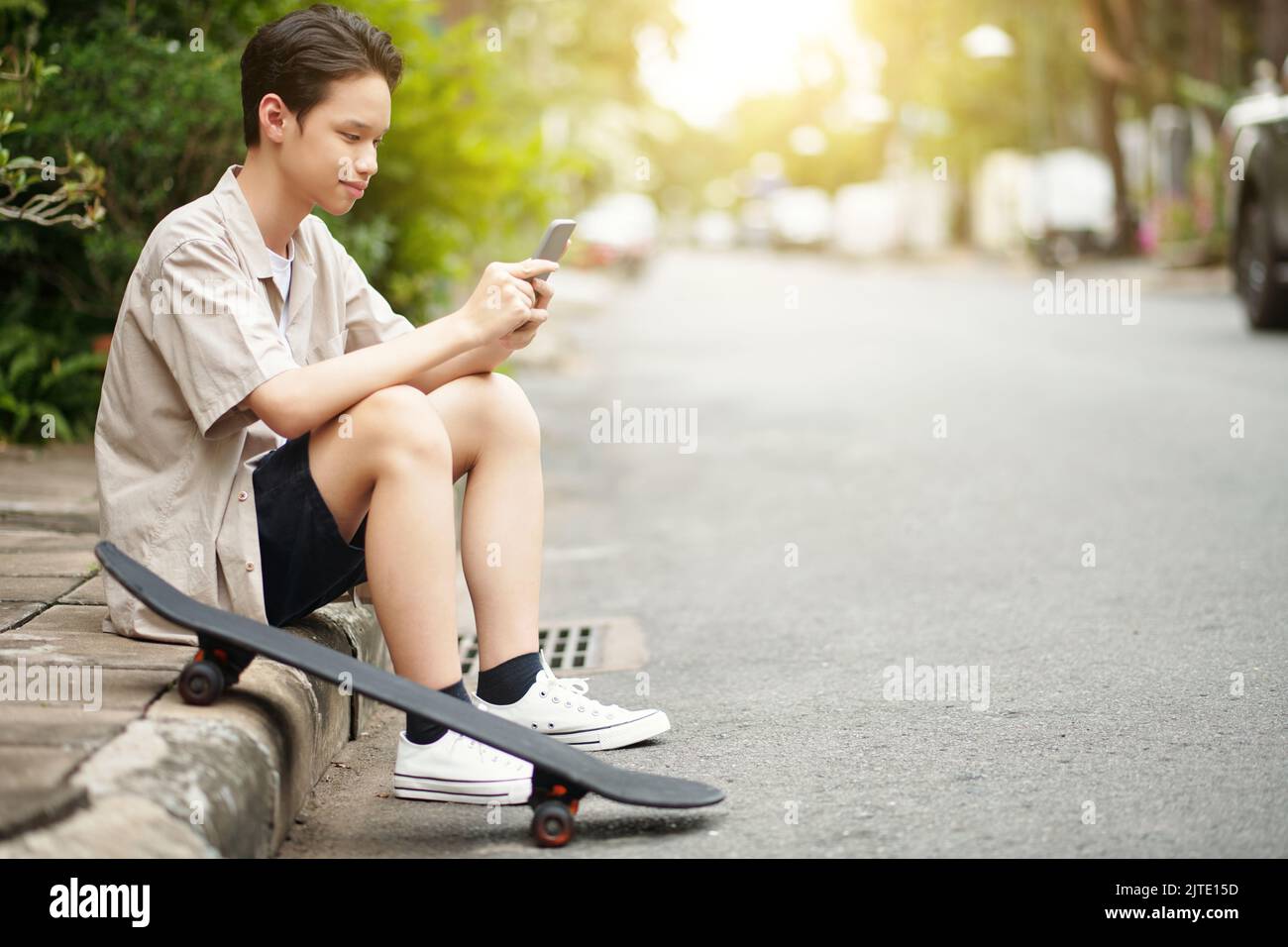Positive teenage boy sitting on pavement next to skateboard and ... Positive teenage boy sitting on pavement next to skateboard and ...