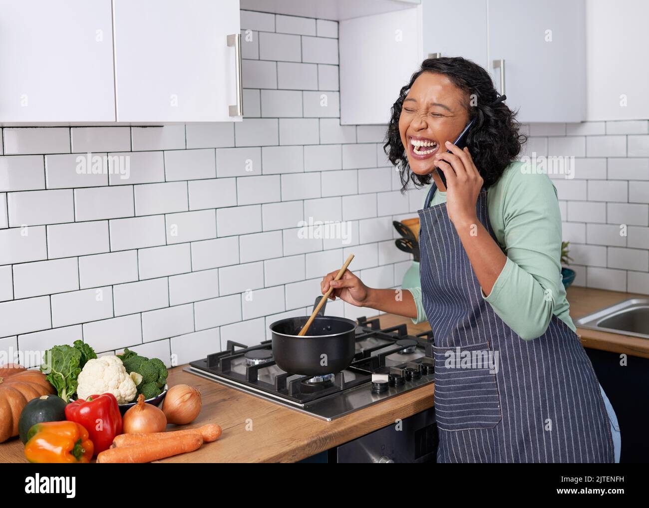 A young multi-racial woman laughs on a phone call while making dinner ...