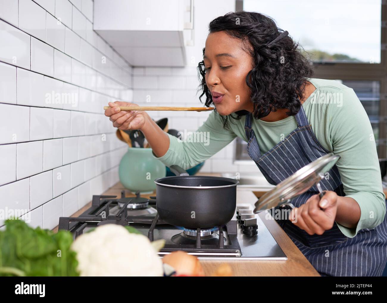 A young multi-racial woman smells and tastes her cooking on the stove ...