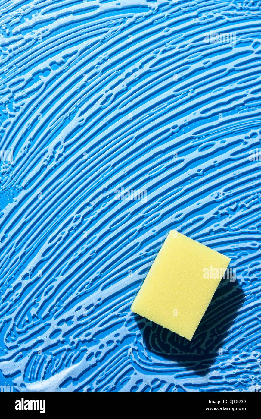 Yellow dishwashing sponge on a blue glass surface covered with soap ...