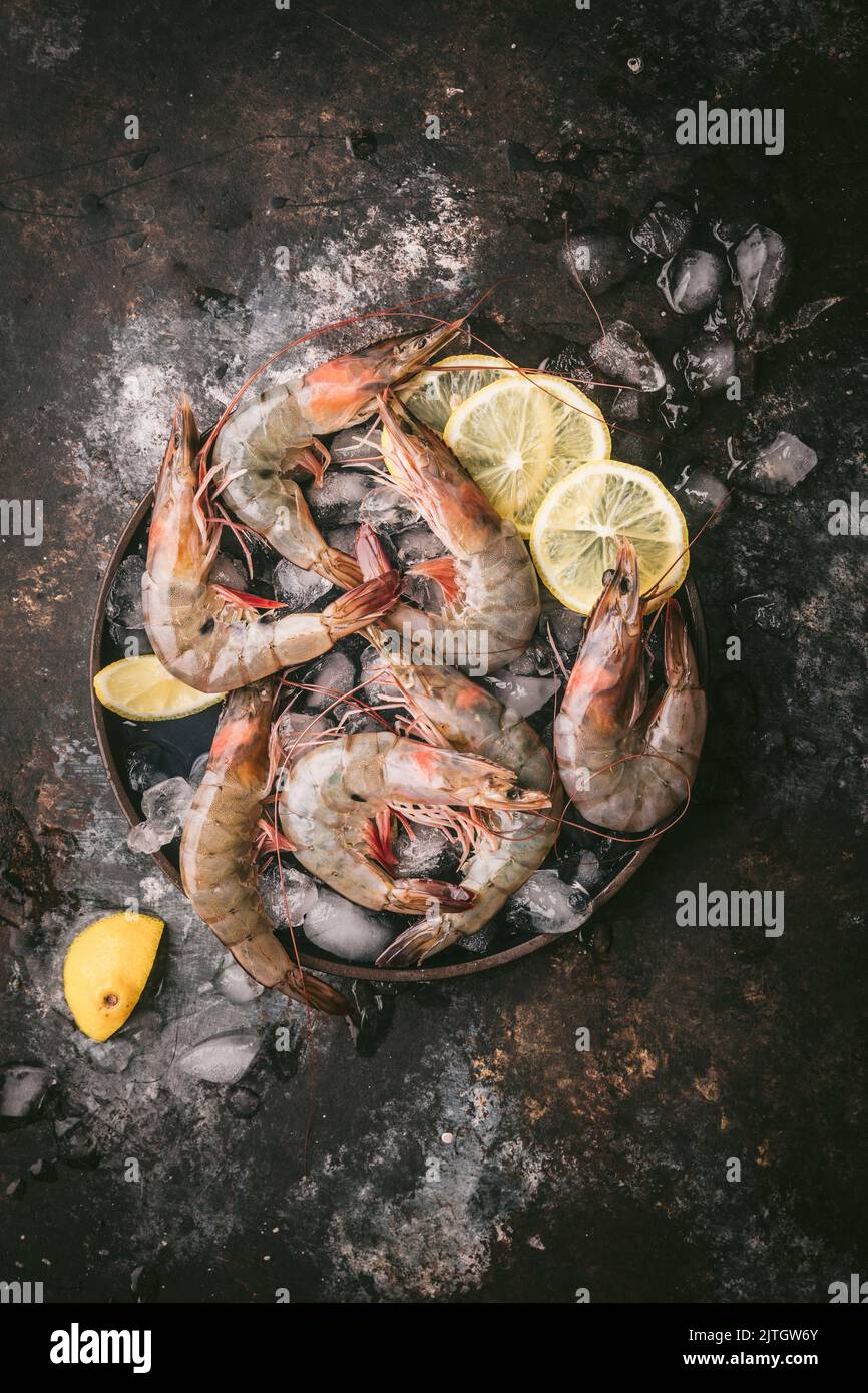 Bowl of raw head-on shrimp on ice with sliced lemons on dark background ...