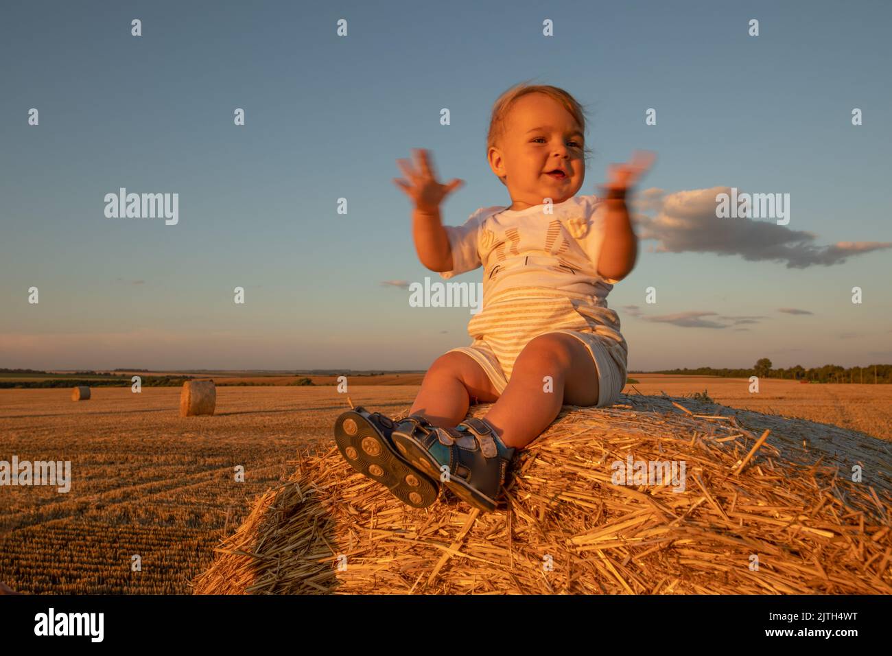 Little cheerful boy, sitting on a big round bale of straw, against the ...