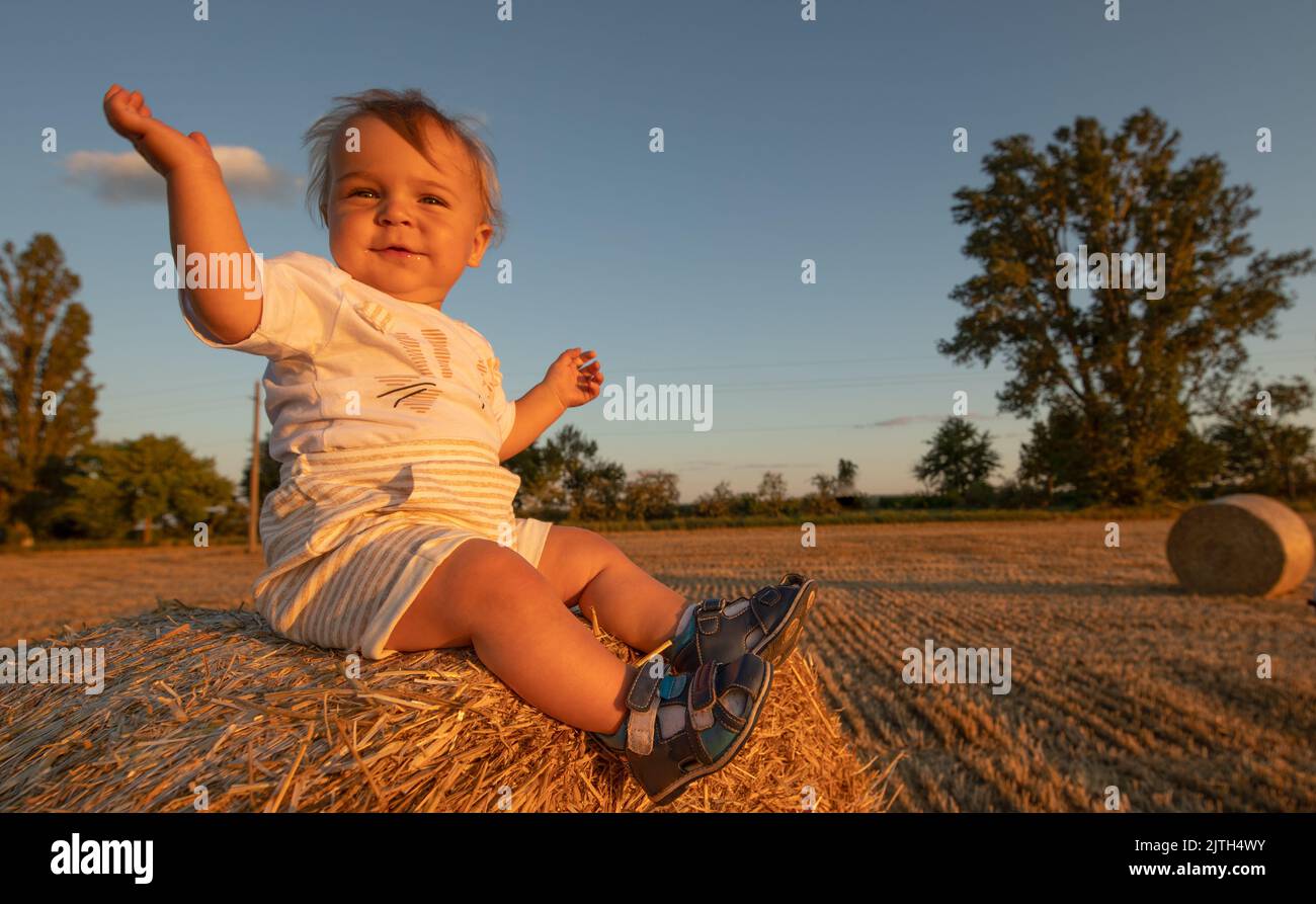 Little cheerful boy, sitting on a big round bale of straw, against the ...