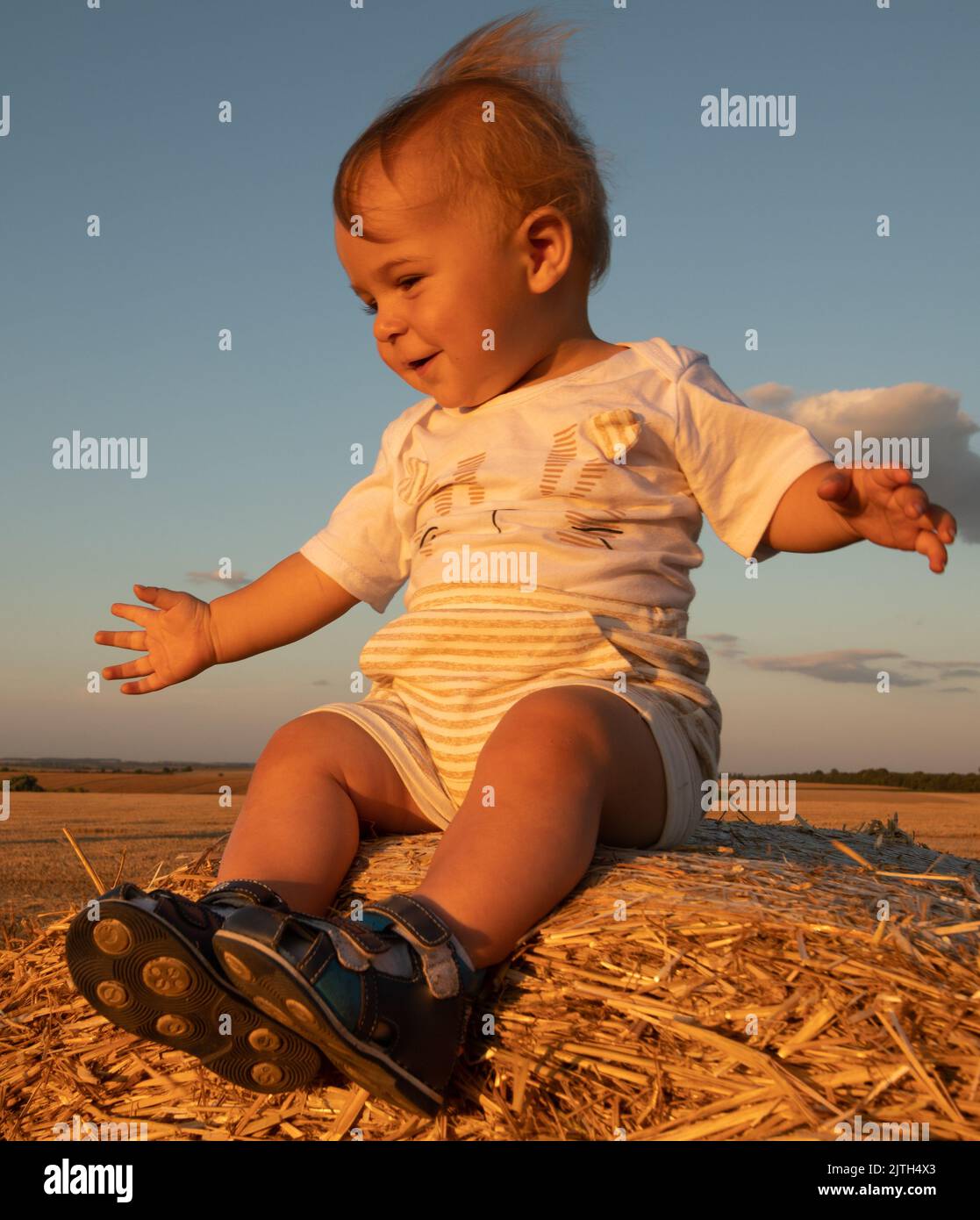 Little cheerful boy, sitting on a big round bale of straw, against the ...