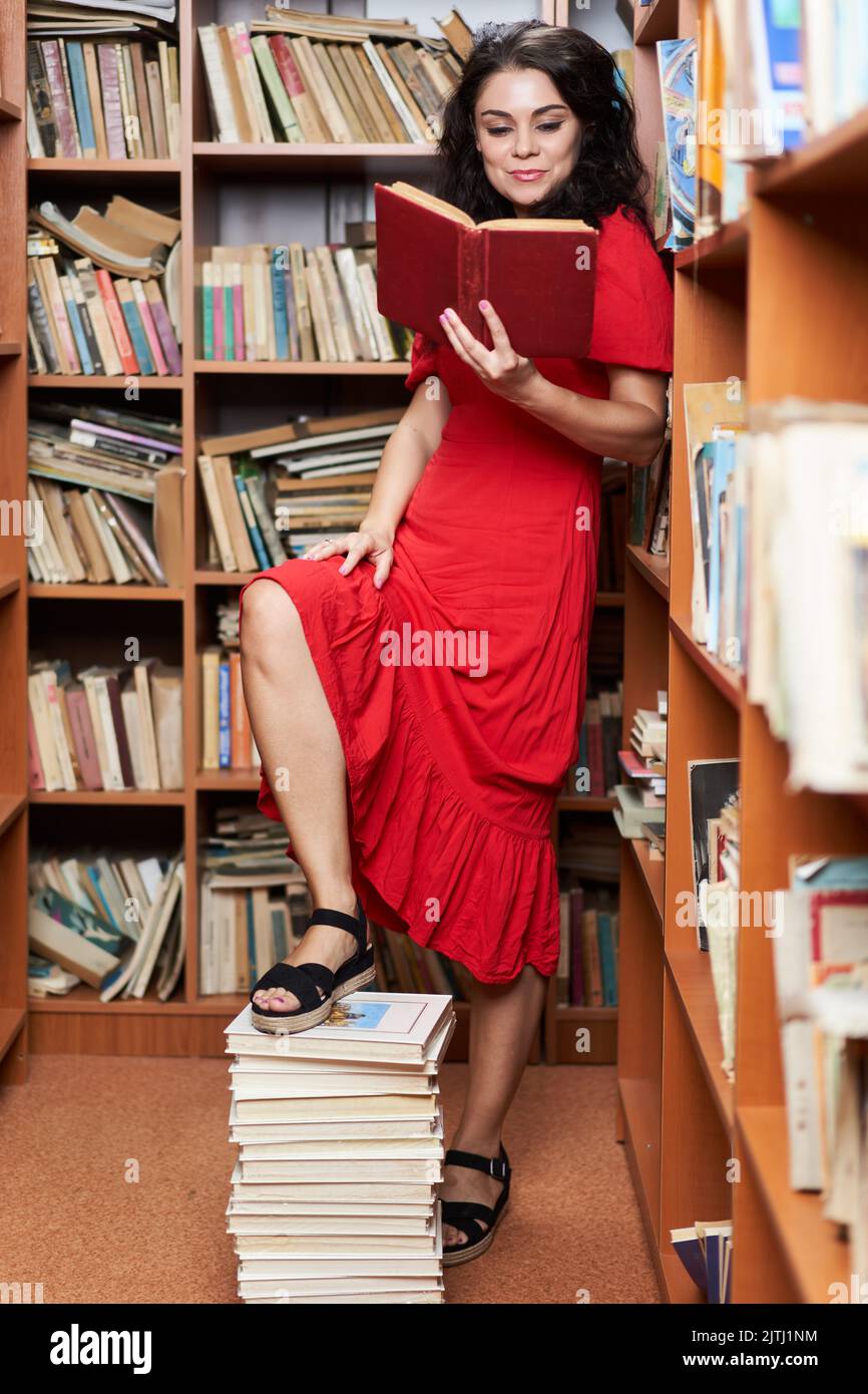 Beautiful woman in red dress in a library sitting on a stack of books ...