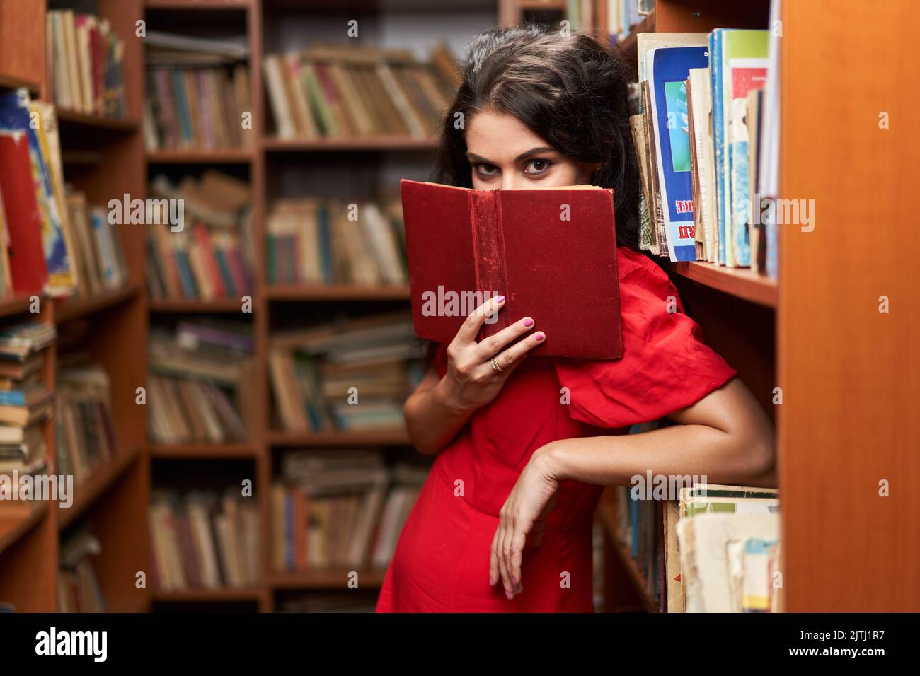 Beautiful woman in red dress in a library, hiding behind an old book ...