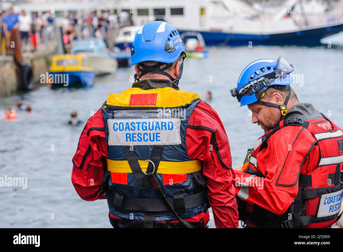 Two men from HM Coastguard Service watch as people swim in a harbour ...