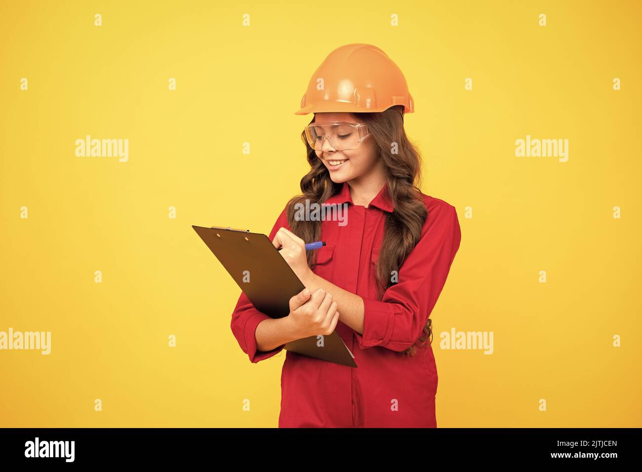 cheerful child girl in protective hard hat and glasses hold folder with ...