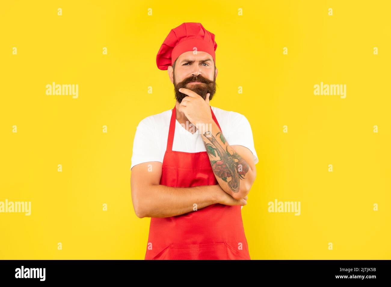 Pensive man in red toque and apron stroking chin yellow background ...