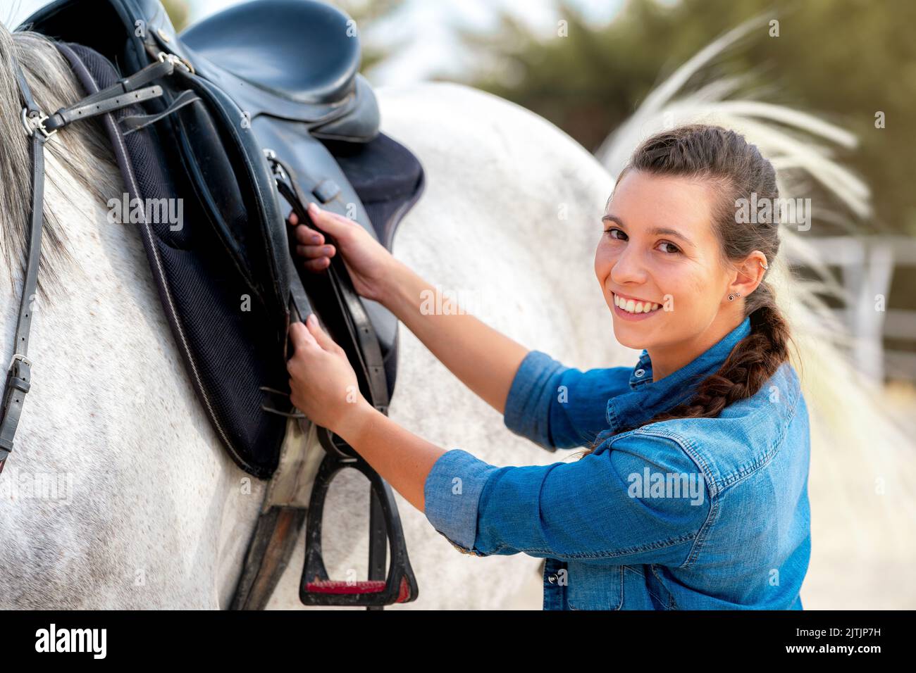 Side view of a woman smiling and putting a saddle on a white horse in a ...